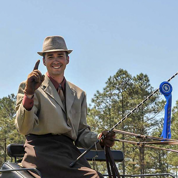 Joe Yoder sits atop a carriage and points a finger to the sky smiling ...